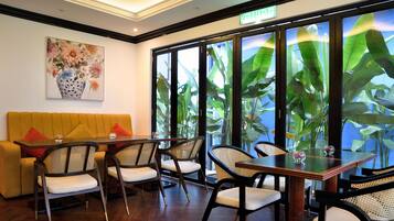 Quarto de hotel com sofá, mesa de jantar, cadeiras e grandes janelas com plantas tropicais.