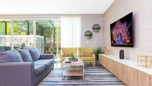 Uma sala de estar moderna com um sofá cinzento, uma poltrona amarela e uma mesa de centro. Uma televisão de ecrã plano está fixada na parede, e existem duas plantas pequenas suspensas na parede.