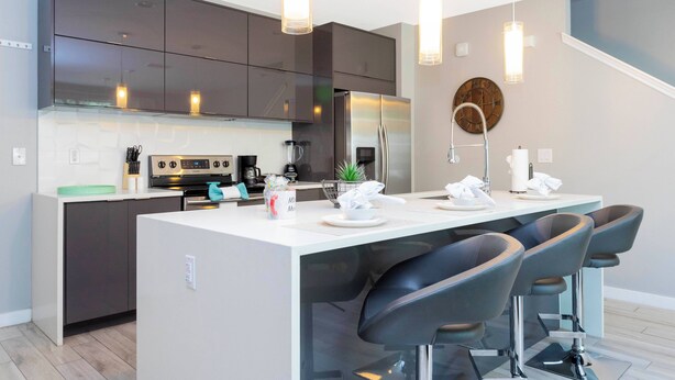 Uma cozinha moderna com armários escuros, uma ilha central com banquetas, eletrodomésticos de aço inoxidável e iluminação pendente.