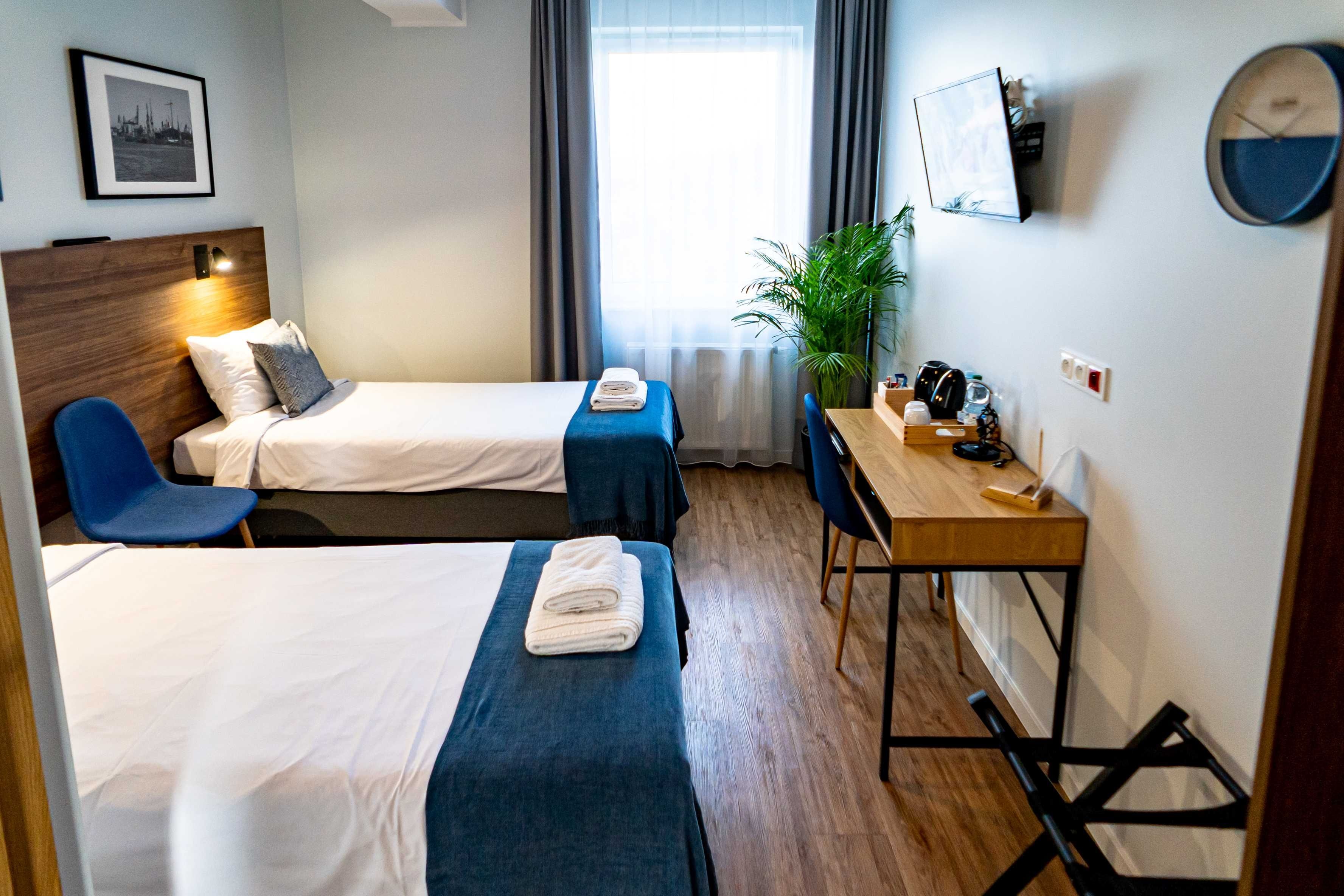 Quarto de hotel com duas camas, uma escrivaninha, uma cadeira, uma planta e um relógio na parede.