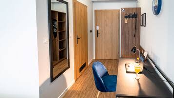 Quarto pequeno com escrivaninha, cadeira e espelho. Há uma porta com uma placa e um relógio de parede.