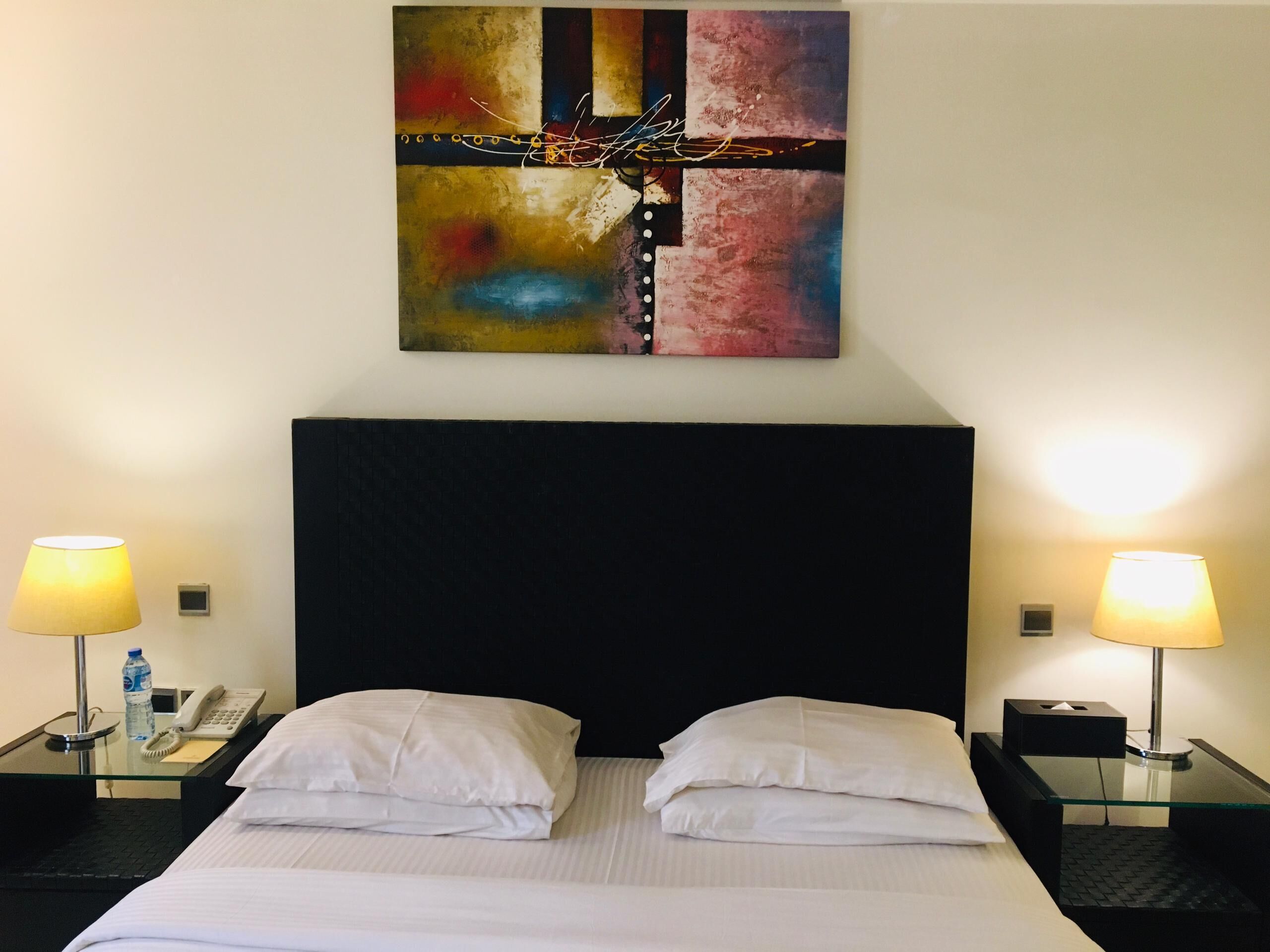 Quarto de hotel com uma cama grande, duas mesas de cabeceira, uma pintura abstrata moderna e duas luminárias.