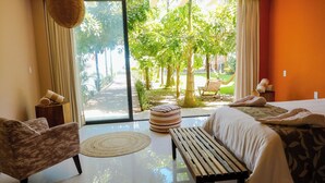 Quarto de hotel moderno com uma grande porta de vidro deslizante que dá para um jardim, uma cama com colcha estampada, um banco de madeira e um banquinho redondo.