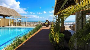 Um deck de madeira que leva a uma piscina com uma estrutura de telhado de palha e uma estátua ao fundo.