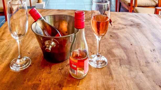 Uma mesa de madeira com uma garrafa de vinho, uma taça de vinho e um balde com abridor de garrafa. A mesa está montada numa sala com cadeiras de madeira e vista para o mar através de grandes janelas.
