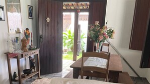 Quarto com cama, mesa de madeira, cadeira, pequena mesa de apoio com objetos decorativos e vista para o exterior.
