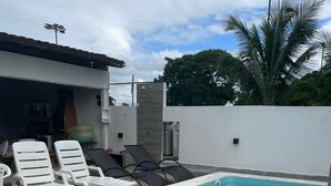 Piscina retangular com borda ladrilhada, cercada por cadeiras de sol brancas e pretas.