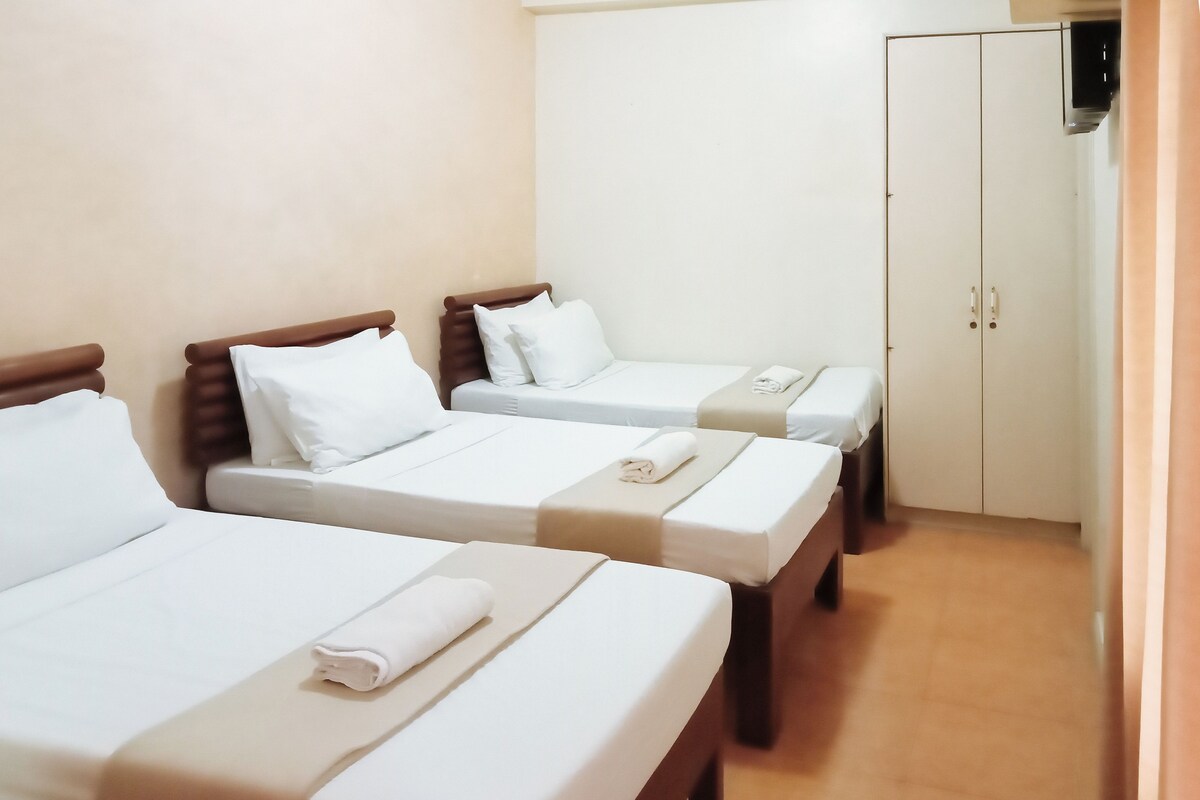 Quarto de hotel com três camas de solteiro, cada uma com roupa de cama e almofadas brancas, e um guarda-roupa com duas portas.