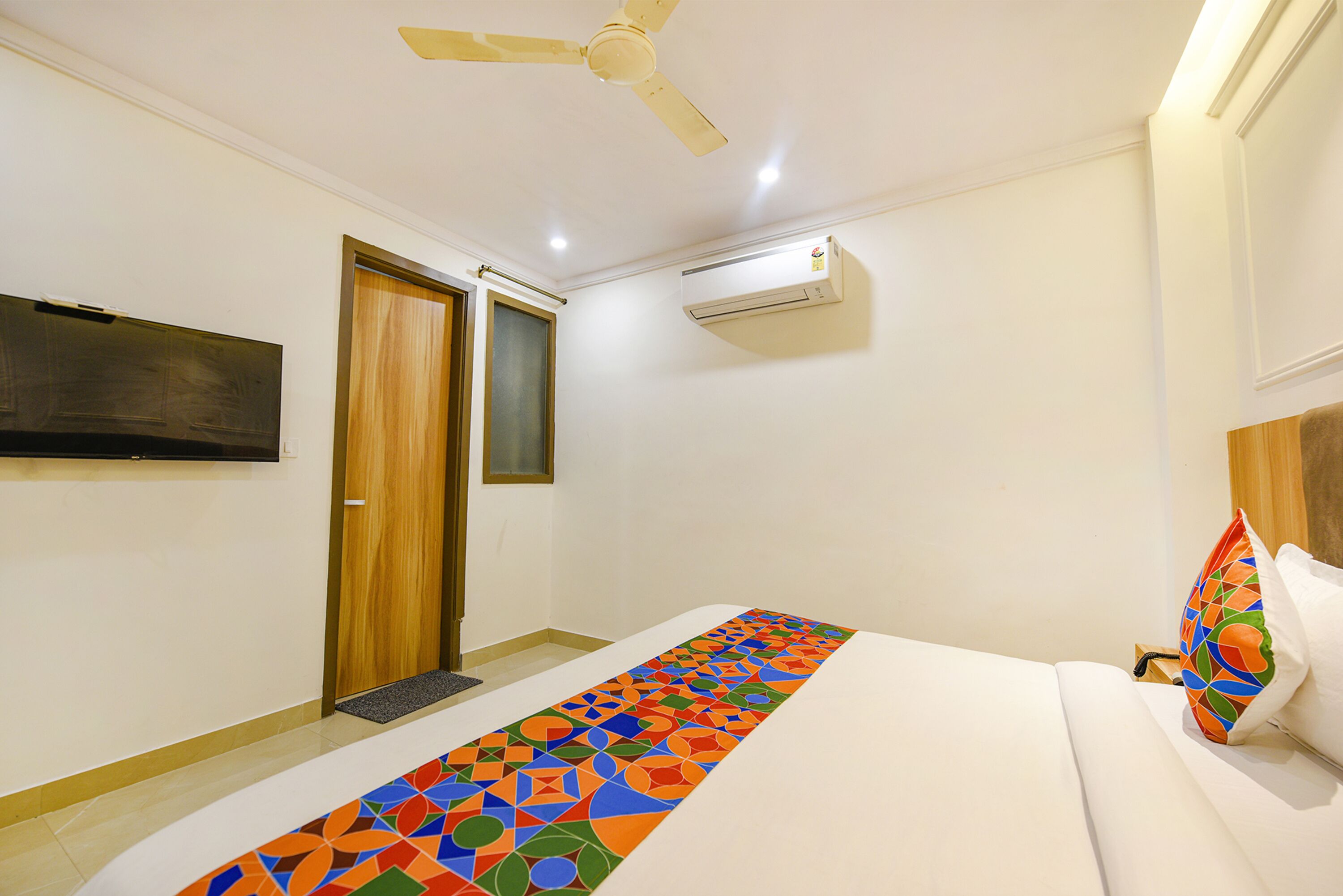Quarto de hotel com cama, ventilador de teto, aparelho de ar condicionado, televisão fixada na parede e um corredor com padrão geométrico colorido.
