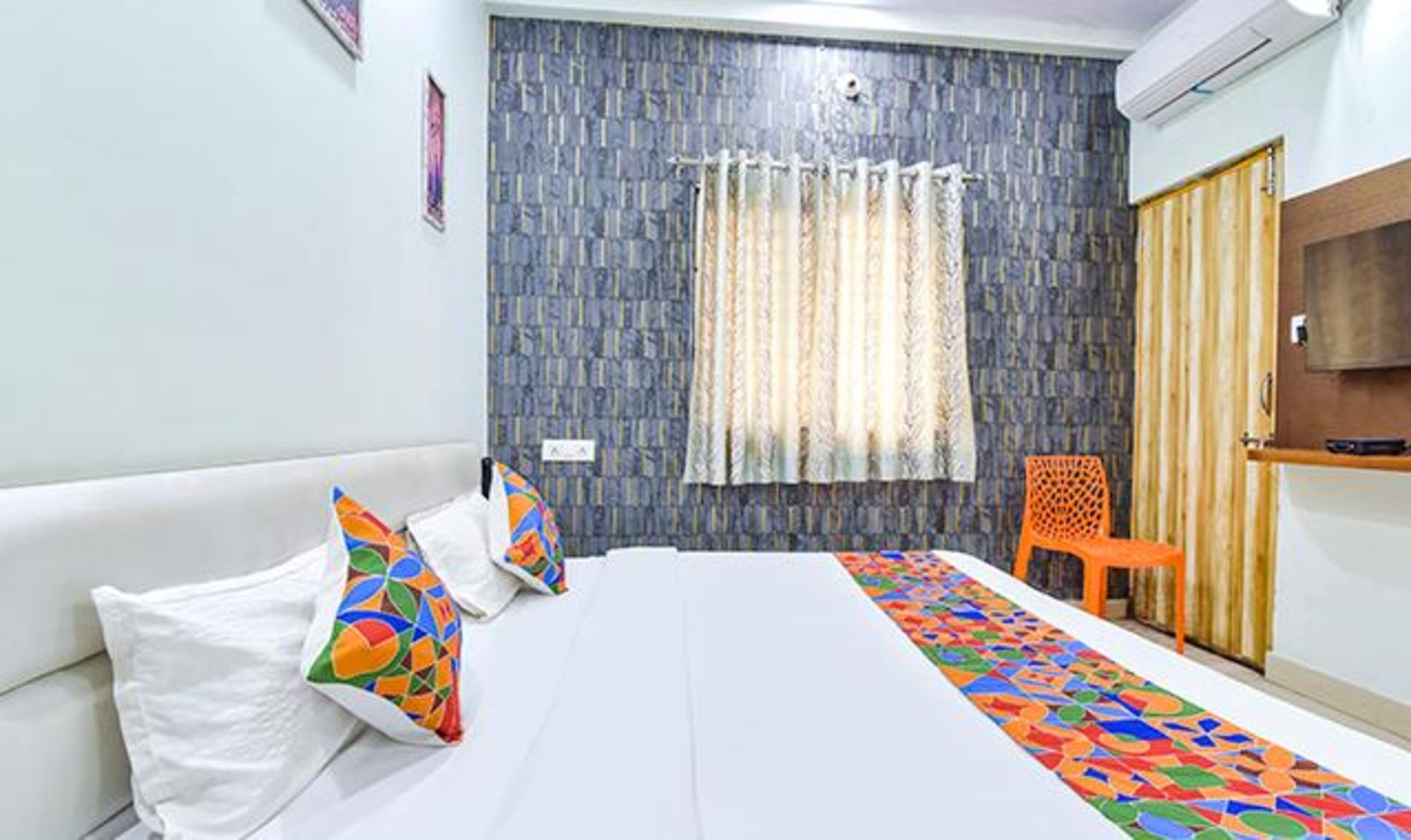 Quarto de hotel com uma cama, almofadas brancas, um corredor de cama colorido, uma janela com cortinas, uma porta de madeira e uma cadeira.