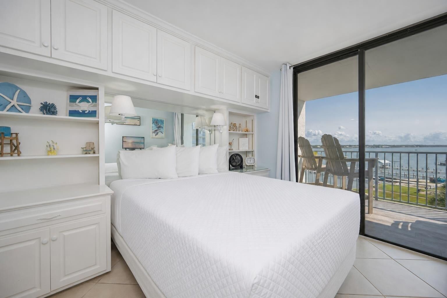 Quarto com uma cama grande, armários brancos, obras de arte e uma varanda com cadeiras.