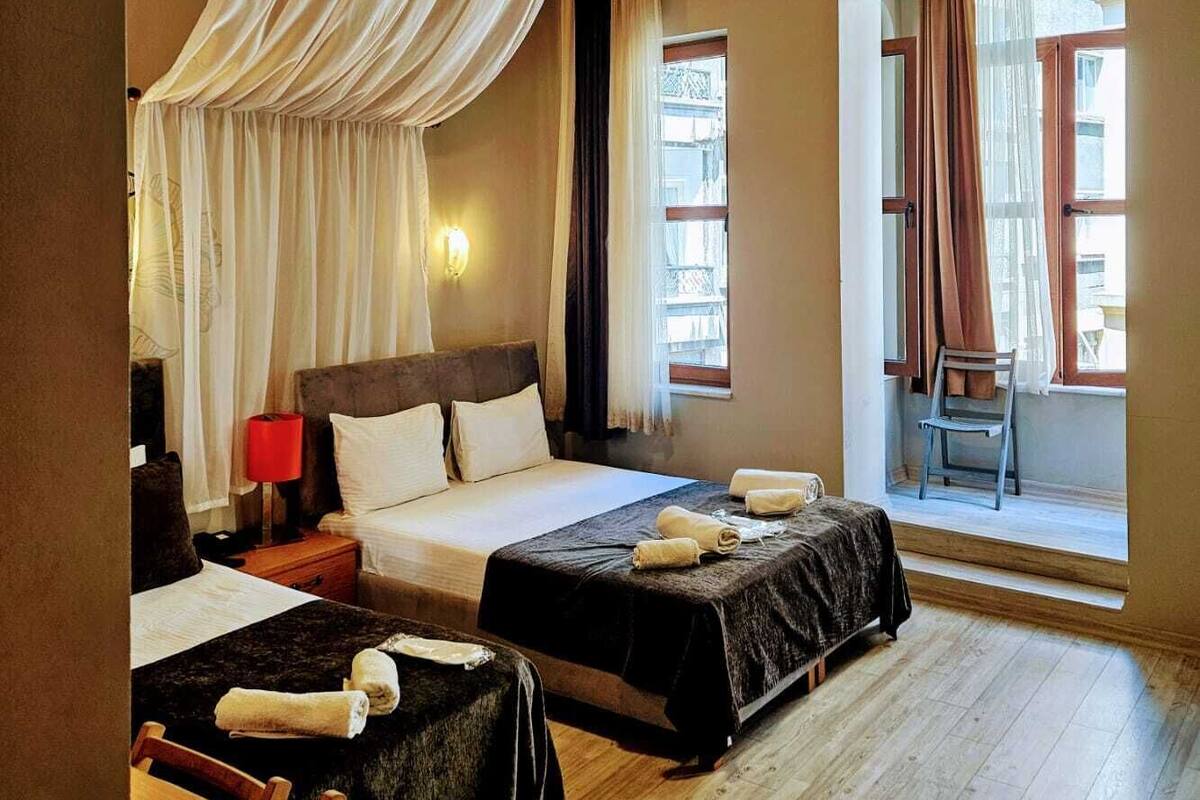 Quarto de hotel com duas camas, teto de madeira, janela com cortinas, uma cadeira e mesa de cabeceira com abajur.