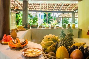 Uma mesa com frutas variadas, em um ambiente tropical com teto de azulejos e plantas em vasos.