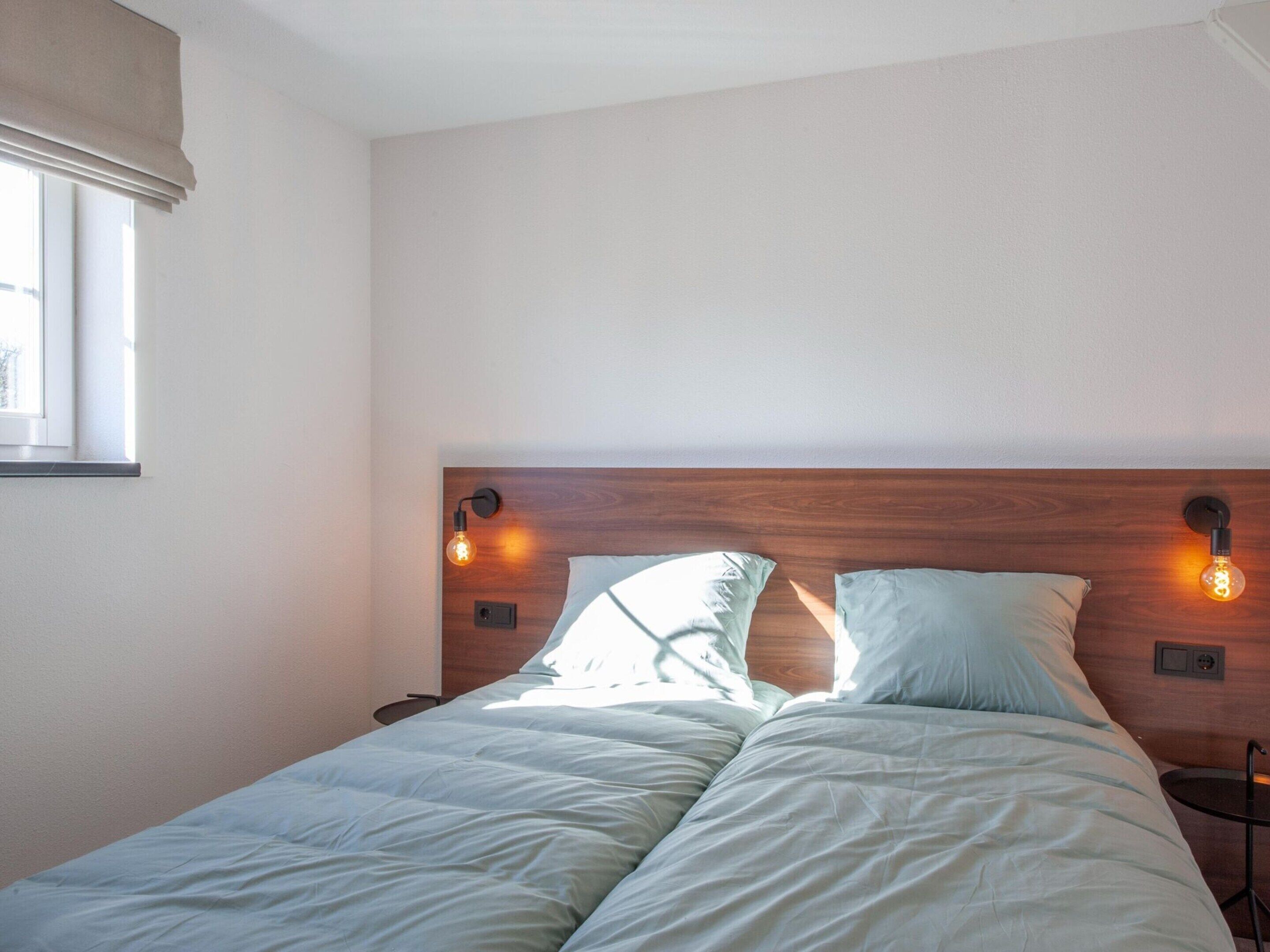Uma cama de casal com cabeceira de madeira, duas luminárias de cabeceira e uma janela com cortinas.