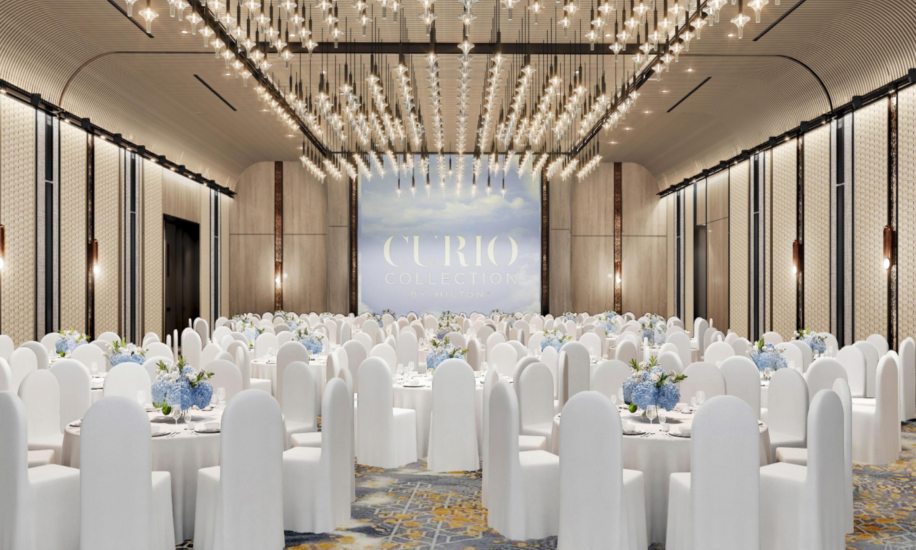 Uma grande sala de conferências com fileiras de mesas redondas, cadeiras brancas e um painel de fundo com o texto 'CURIO COLLECTION BY HILTON'.