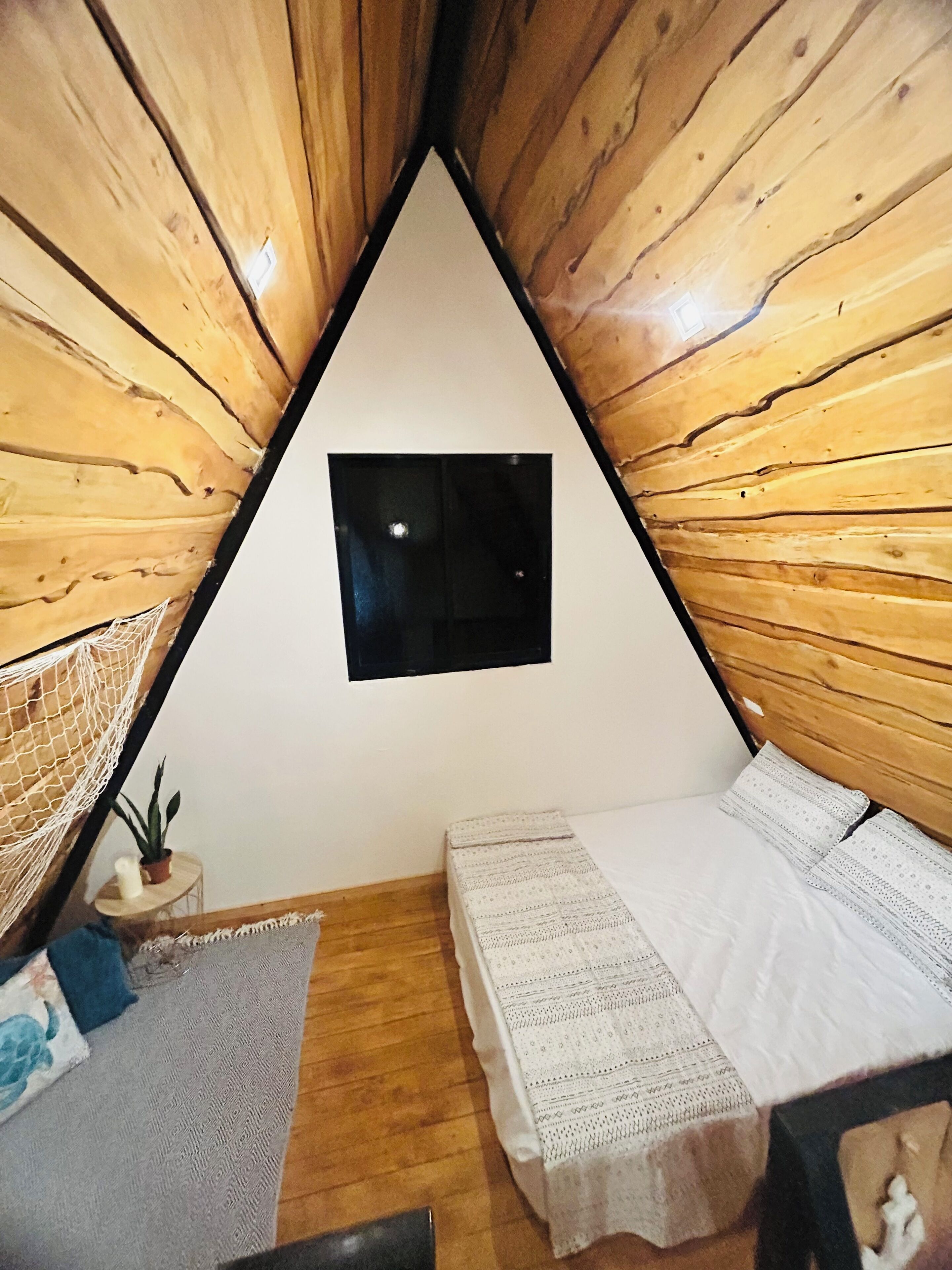 Um quarto triangular com paredes de madeira, uma pequena janela preta, duas camas com lençóis brancos, uma mesinha com uma planta e uma rede.