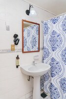 Um banheiro com pia branca, espelho, cortina de chuveiro com padrão floral azul e luminária fixa na parede.