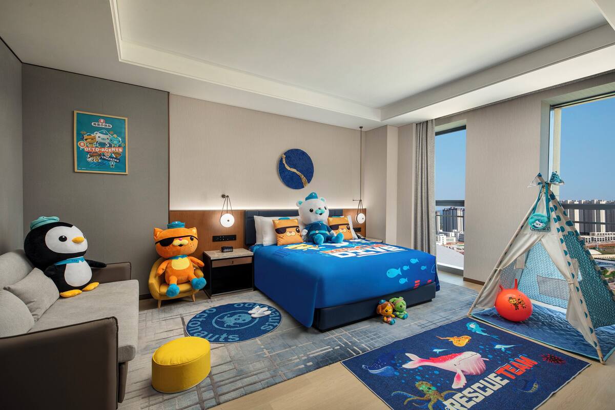 Quarto de hotel com cama, sofá, tenda infantil e uma grande janela com vista para a cidade.
