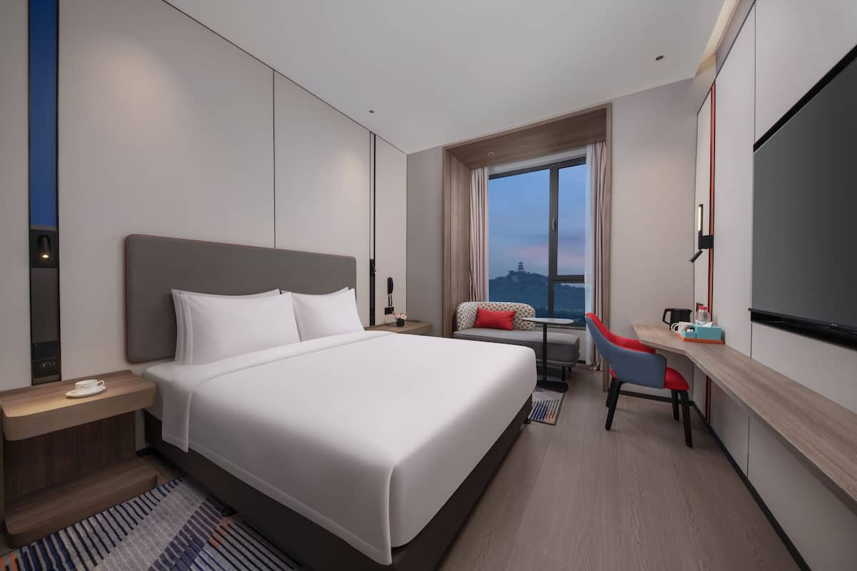 Quarto de hotel com uma cama grande, uma escrivaninha com cadeira, televisão e janela com vista.