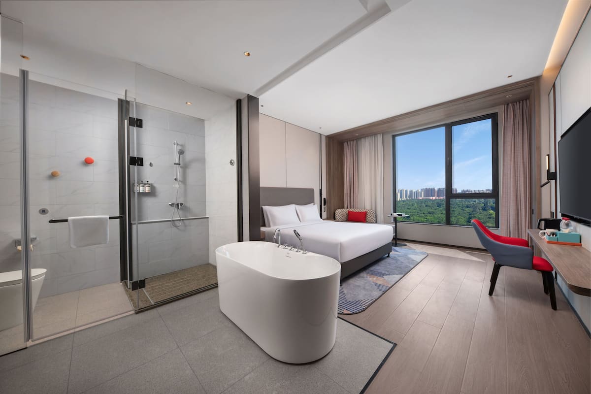 Quarto de hotel moderno com chuveiro em cabine de vidro, uma banheira grande, cama com vista para a cidade e uma mesa de trabalho com cadeira.