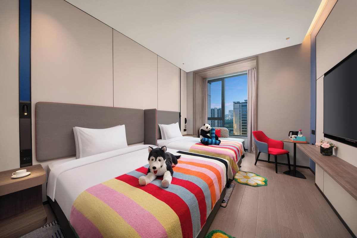 Quarto de hotel com duas camas, televisão, secretária e janela com vista para a cidade.