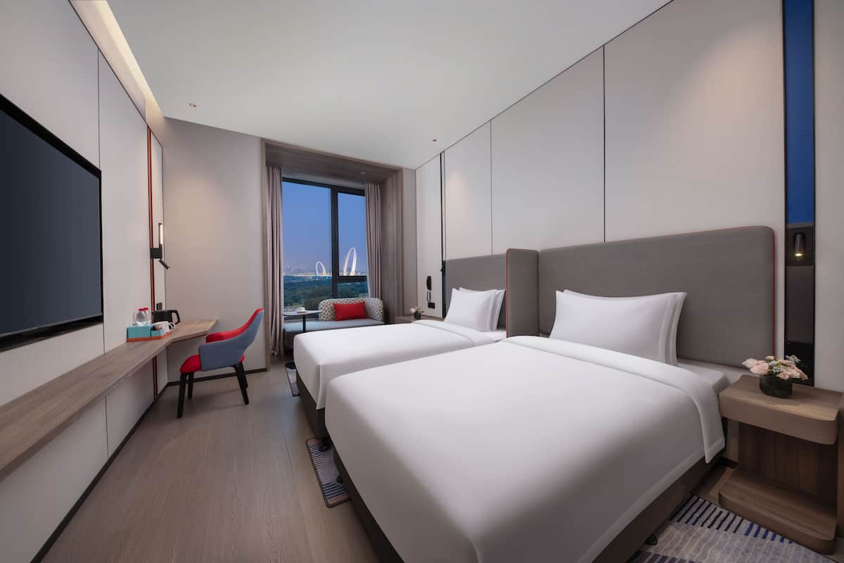 Quarto de hotel com duas camas, uma mesa com cadeira, televisão e vista para uma ponte.