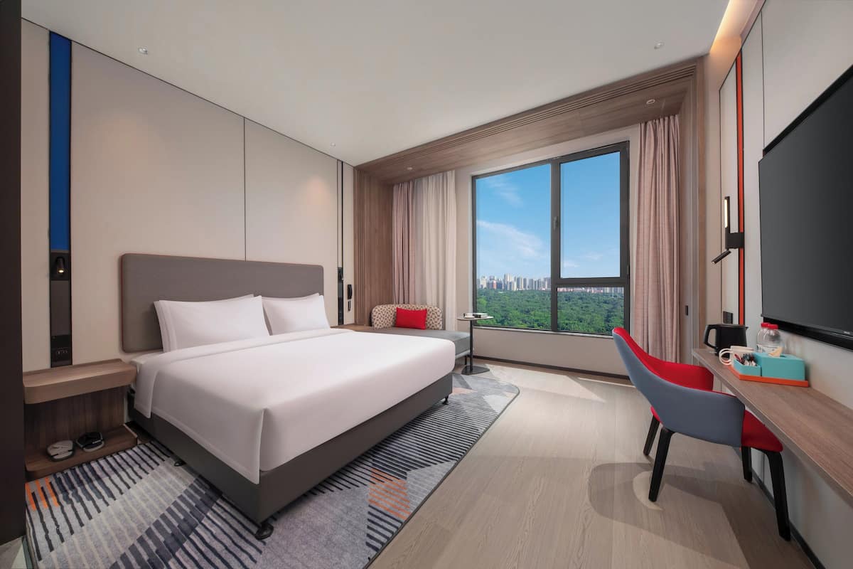 Quarto de hotel moderno com uma cama grande, uma escrivaninha com cadeira, televisão e vista para a cidade pela janela.
