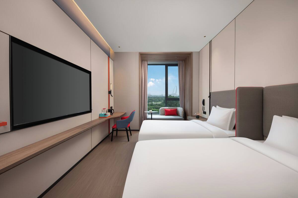 Quarto de hotel moderno com uma grande televisão de tela plana, uma mesa com cadeira, duas camas e vista para a paisagem urbana através de amplas janelas.