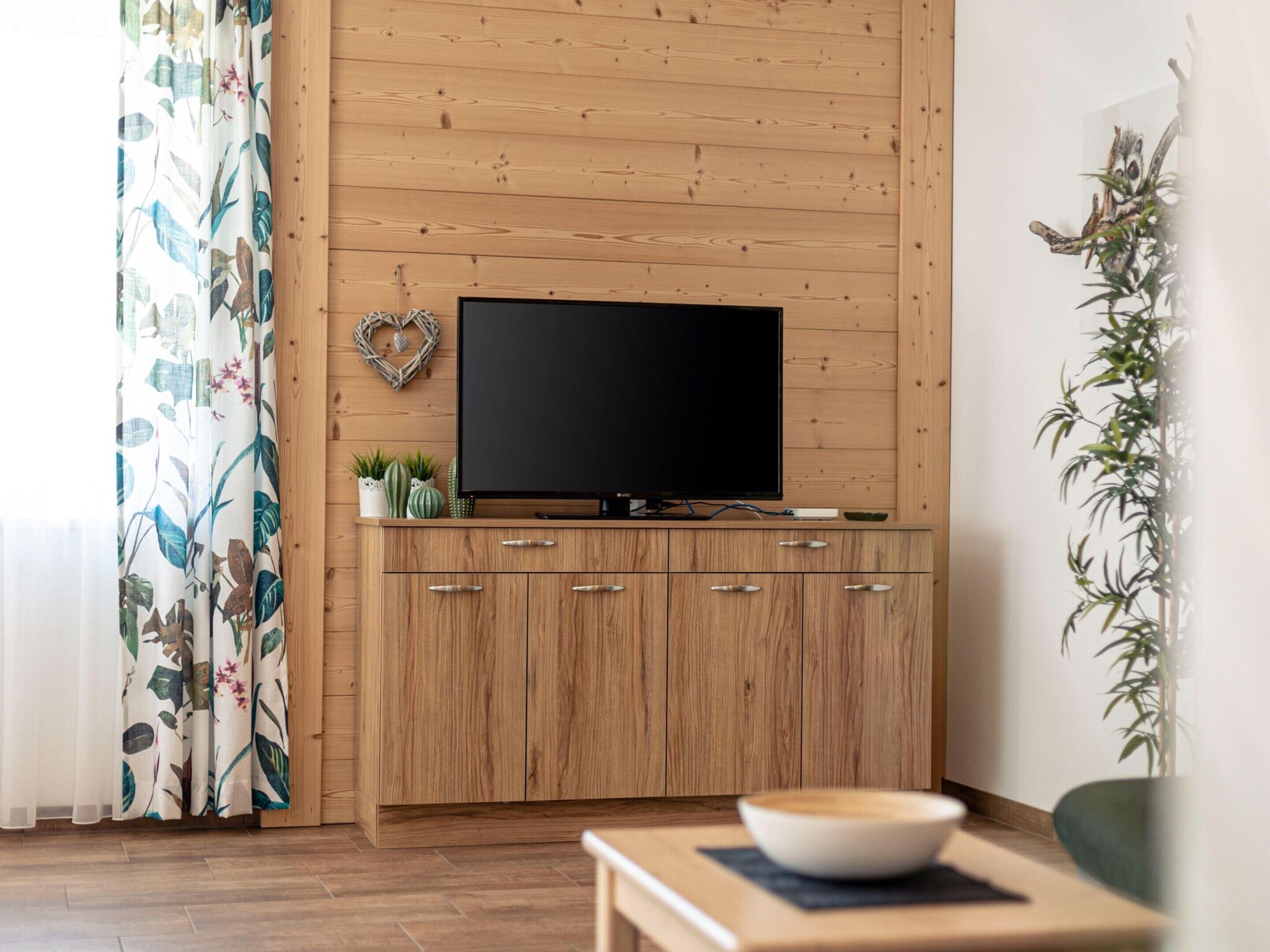 Uma sala de estar com um armário de madeira, uma TV de tela plana, um enfeite em formato de coração e uma planta.
