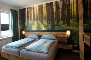 Uma cama de casal com um mural da floresta na parede, uma janela com cortinas e uma mesa de cabeceira com abajur.