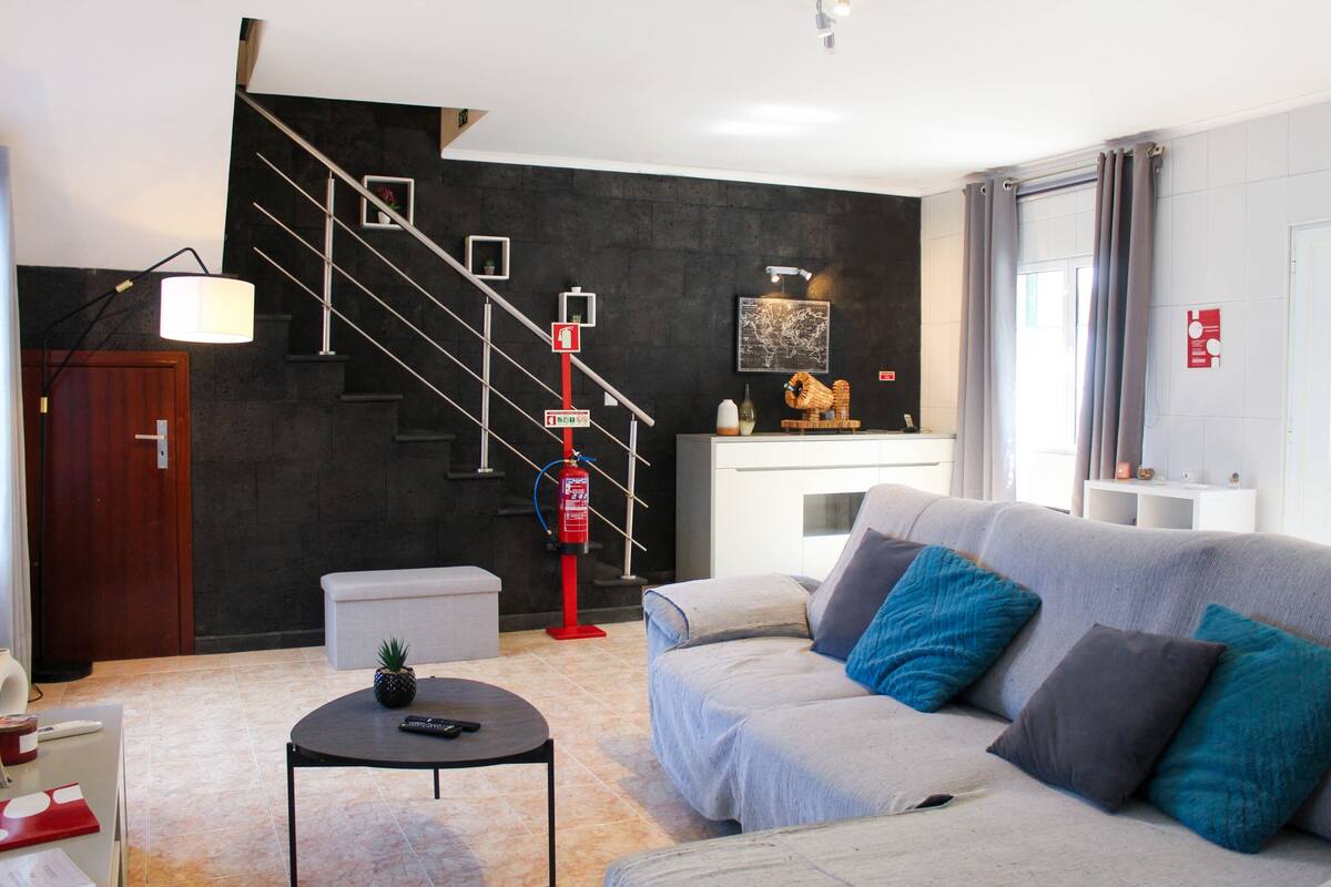 Uma sala de estar moderna com um sofá cinza, uma mesa redonda pequena e uma escada com um extintor de incêndio vermelho.