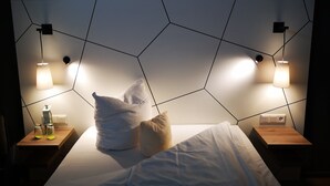 Quarto de hotel moderno com cabeceira estampada em padrão geométrico, duas luminárias de cabeceira, mesa de cabeceira de madeira e uma garrafa de água.