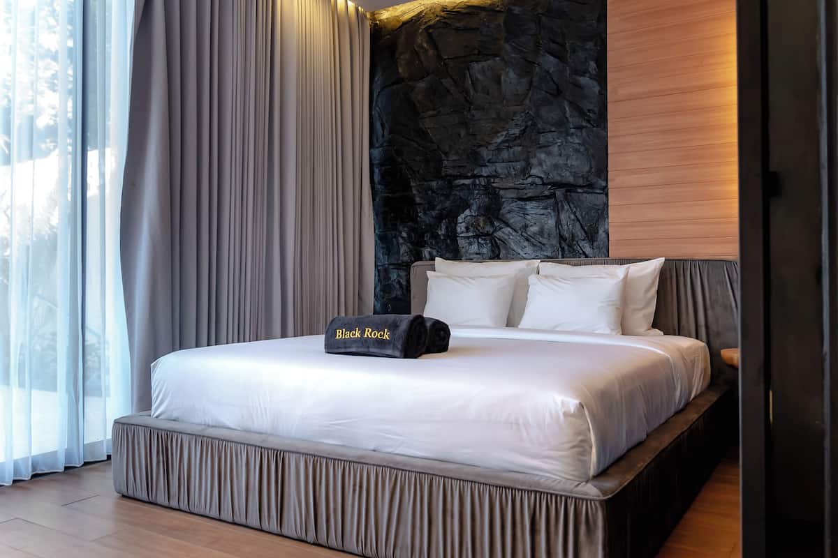 Quarto de hotel moderno com uma cama grande, parede de destaque em pedra e vista para o exterior através de cortinas leves.