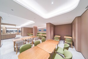 Uma sala de jantar moderna com várias mesas e cadeiras, iluminação embutida no teto e vista para a cozinha ao fundo.