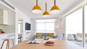 Uma sala de estar moderna com uma mesa de jantar de madeira, cadeiras brancas e um sofá com almofadas amarelas. Três luminárias pendentes estão sobre a mesa, e uma janela grande com cortinas leves.