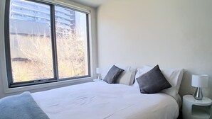 Quarto de hotel moderno com uma cama grande, roupa de cama branca e vista para um edifício pela janela.