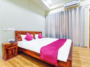 Cama bem arrumada com cabeceira de madeira, mesa de cabeceira com telefone, e quarto iluminado com claraboia e cortinas.