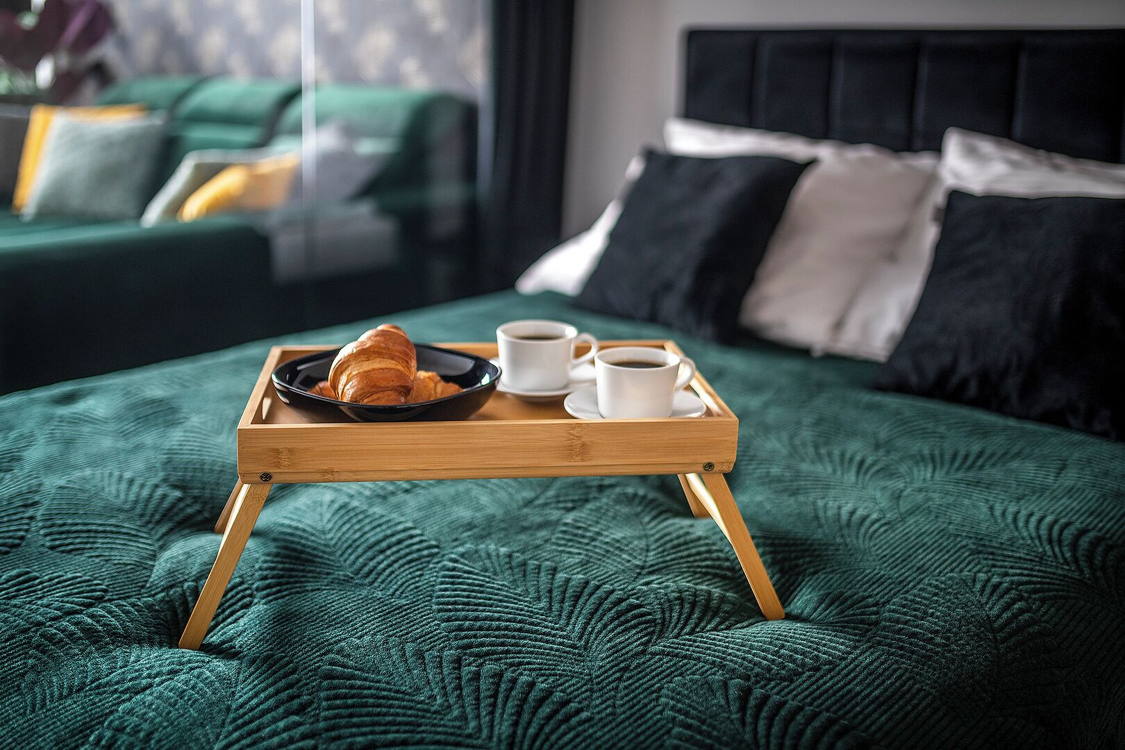 Cama com um edredom verde texturizado, uma bandeja de madeira com croissants e xícaras de café, e ao fundo, um sofá verde e uma janela.