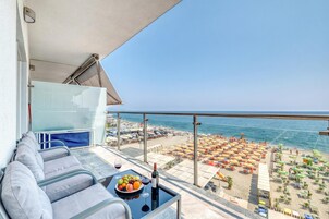 Varanda com sofá, mesa com frutas e vinho, e vista para uma praia com guarda-sóis e cadeiras de praia.