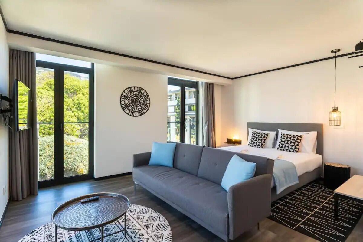 Quarto de hotel moderno com uma cama grande, um sofá cinzento, uma mesa redonda pequena e vista para o verde pelas janelas.