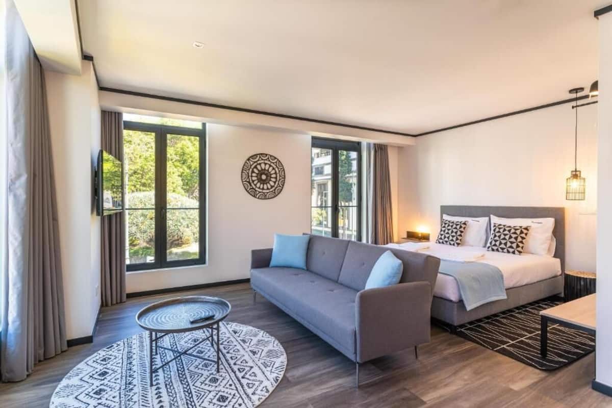 Quarto de hotel moderno com sofá, cama, mesa de centro redonda e janelas amplas com vista para o verde lá fora.