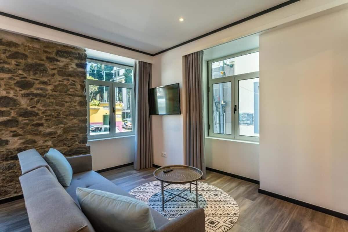 Uma sala de estar moderna com uma parede de destaque em pedra, um sofá cinzento, uma mesa de centro redonda e grandes janelas com cortinas.