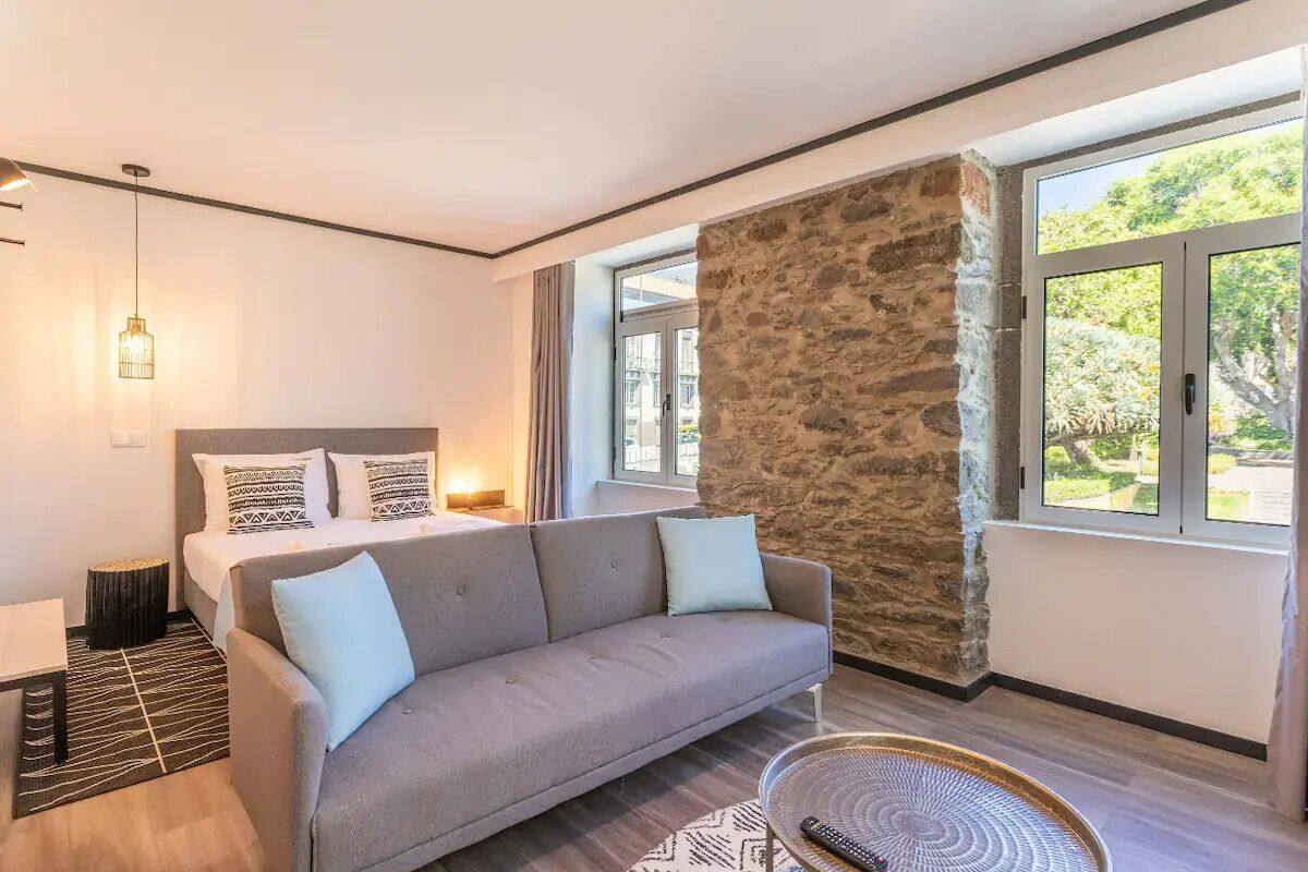 Quarto de hotel moderno com cama, sofá, parede de pedra decorativa e janelas amplas.