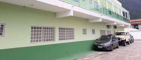 Um edifício com vários andares, fachada verde e branca, varanda de vidro e área de estacionamento com dois carros.