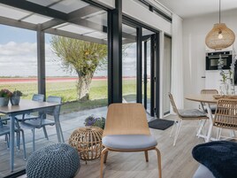 Uma área de refeições ao ar livre moderna com vista para um campo e uma árvore, apresentando uma mesa de madeira, cadeiras azuis e mobiliário de vime.
