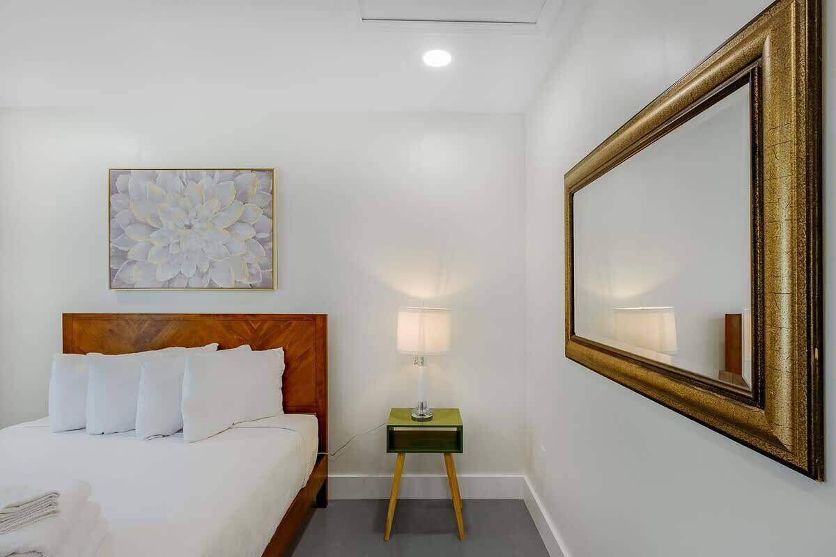 Quarto de hotel com cama, mesa de cabeceira com abajur, um espelho grande e uma obra de arte na parede.
