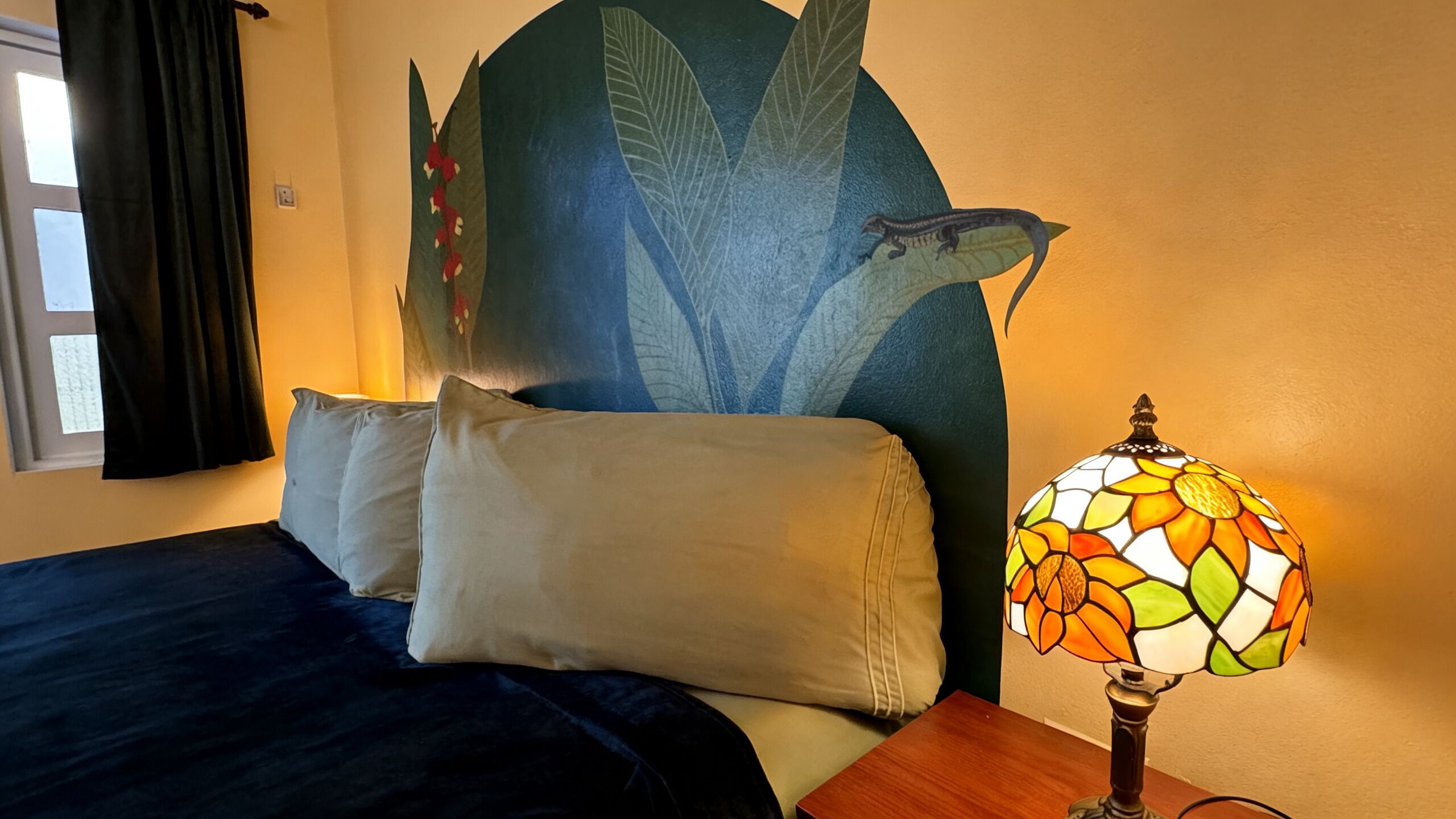 Uma cama com almofadas, uma mesa de cabeceira com abajur e um mural na parede.