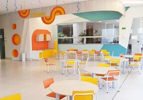 Uma cafeteria vibrante, com mesas e cadeiras redondas, um balcão com cardápio e uma área de recreação com um escorrega.