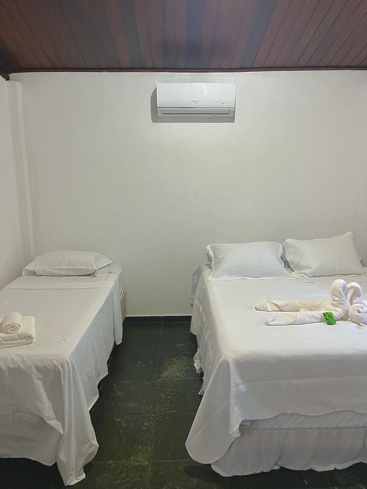 Quarto com duas camas individuais, cada uma com um travesseiro e uma toalha enrolada. Um aparelho de ar condicionado está fixado na parede acima das camas.
