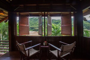 Quarto com duas cadeiras de madeira voltadas para uma janela com portadas abertas, com vista para uma paisagem verde exuberante.